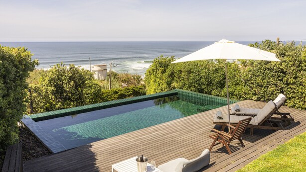 Piscina privativa com vista para o oceano, deck de madeira, cadeiras de descanso e uma mesa com um copo de vinho.