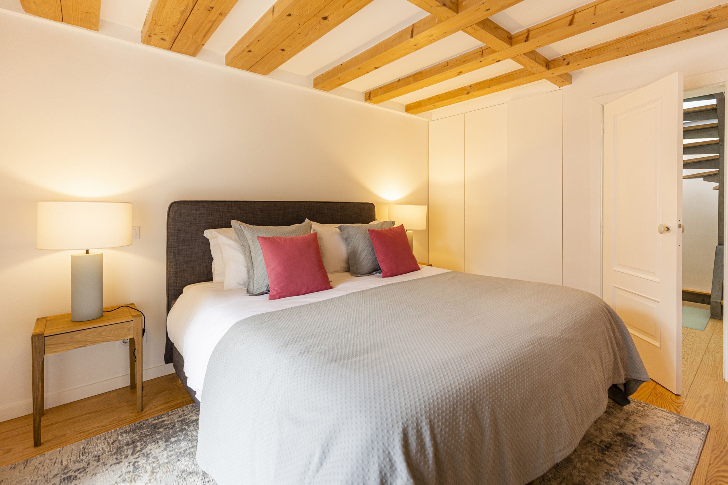 Quarto com cama, luminárias de cabeceira, painel de madeira e porta que dá para uma escada.