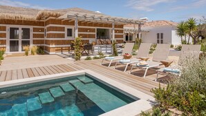 Uma área à beira da piscina com espreguiçadeiras, um deck de madeira e um edifício com revestimento de madeira listrada e telhado de palha.