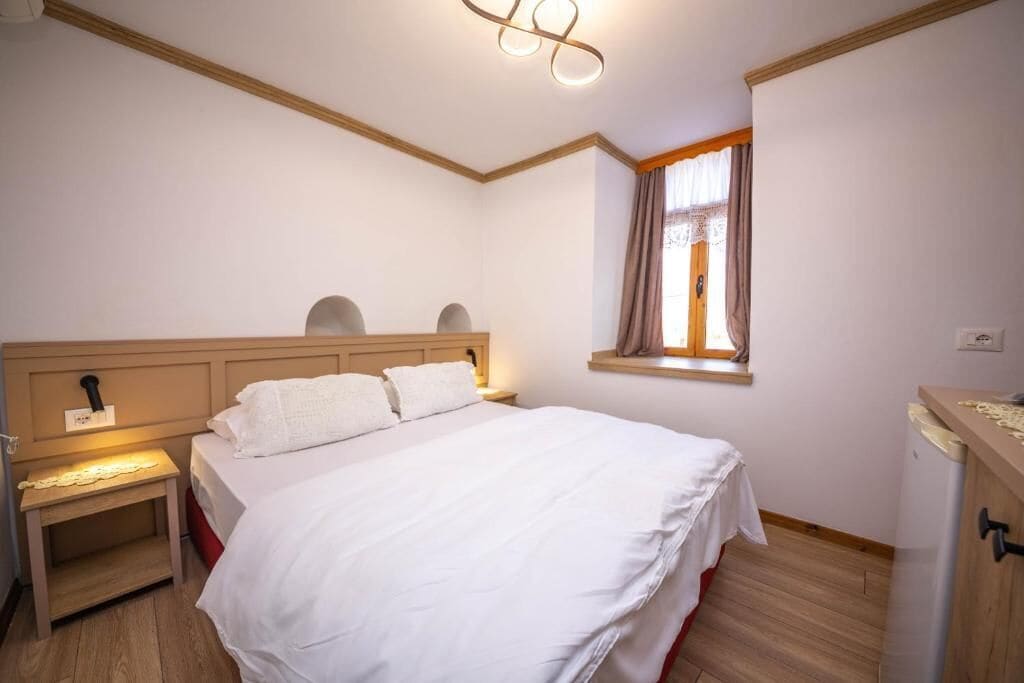 Quarto com cabeceira de madeira, mesa de cabeceira com abajur, janela com cortinas e um pequeno armário.