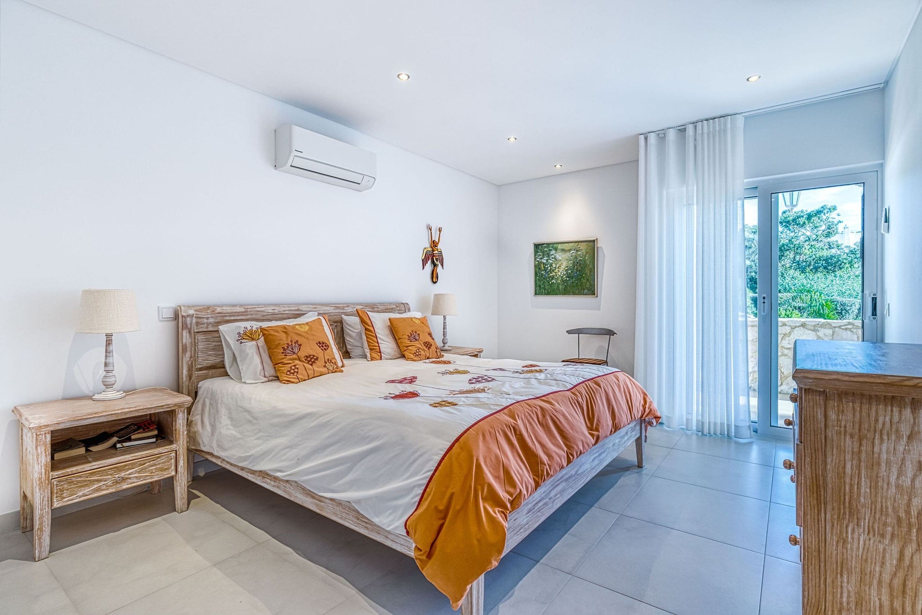 Quarto com uma cama grande, mesa de cabeceira, cômoda de madeira e porta de vidro corrediça para uma varanda.