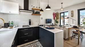 Cozinha moderna com armários brancos, bancadas pretas e uma ilha central. Há uma área de jantar com um sofá e uma mesa de centro. A cozinha possui uma grande janela com vista para a cidade.