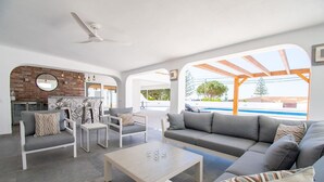 Uma sala de estar moderna com um sofá cinzento, poltronas brancas e uma mesa de centro. Apresenta uma parede de tijolos, um ventilador de teto e vista para uma piscina através de grandes janelas.