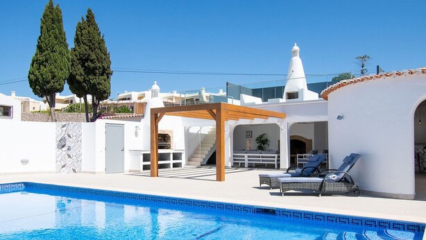 Piscina com espreguiçadeiras e pérgula, cercada por edifícios brancos e ciprestes.