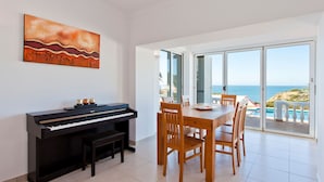 Um piano numa sala com uma mesa de jantar e cadeiras, janelas amplas com vista para o mar e um quadro na parede.