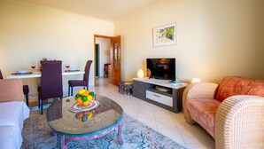 Uma sala de estar com um sofá, uma mesa de centro com frutas, uma televisão e um espaço de jantar com cadeiras e uma mesa.