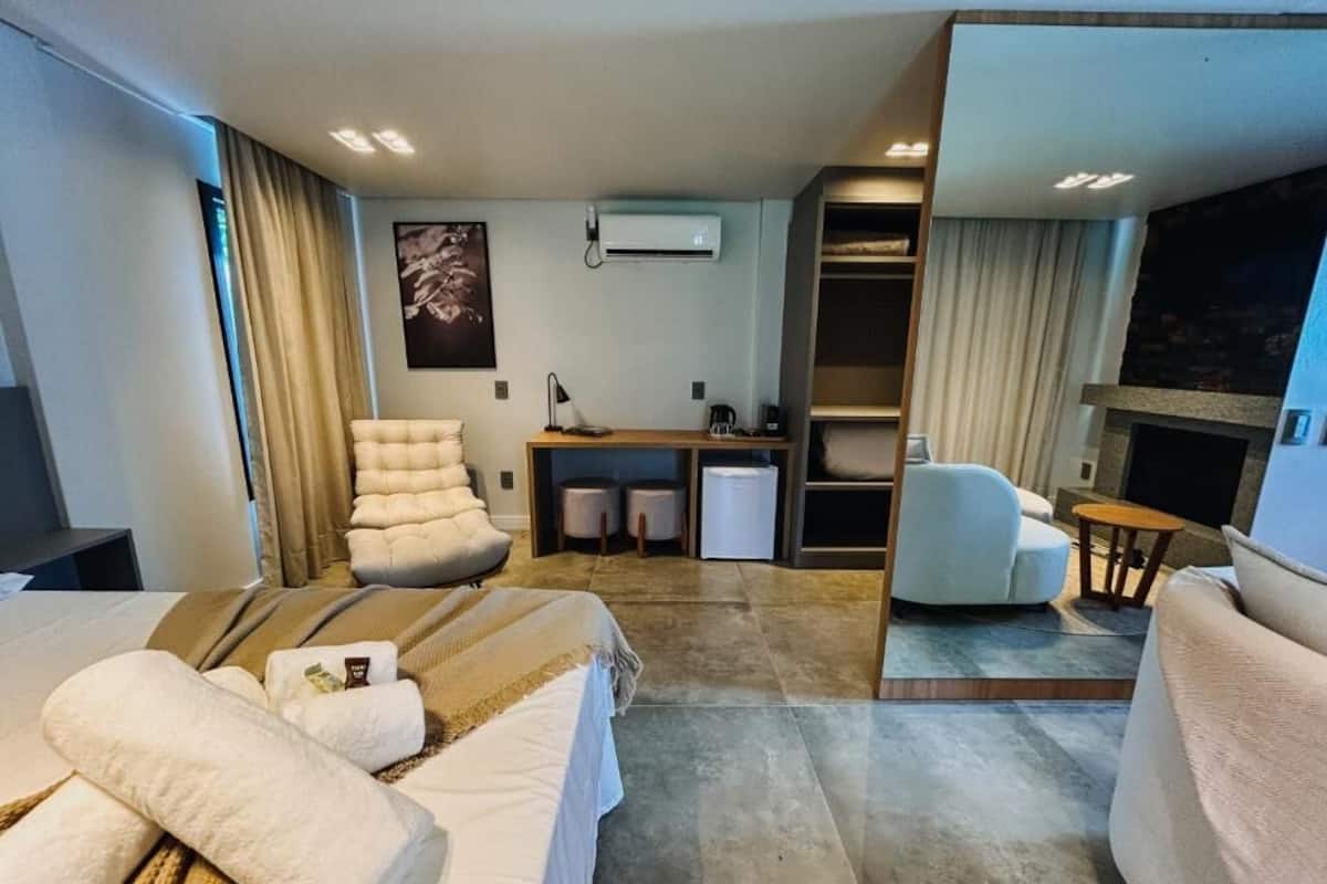 Quarto de hotel moderno com cama, sofá, escrivaninha, cadeira e uma pequena mesa. Possui um guarda-roupa e uma lareira.