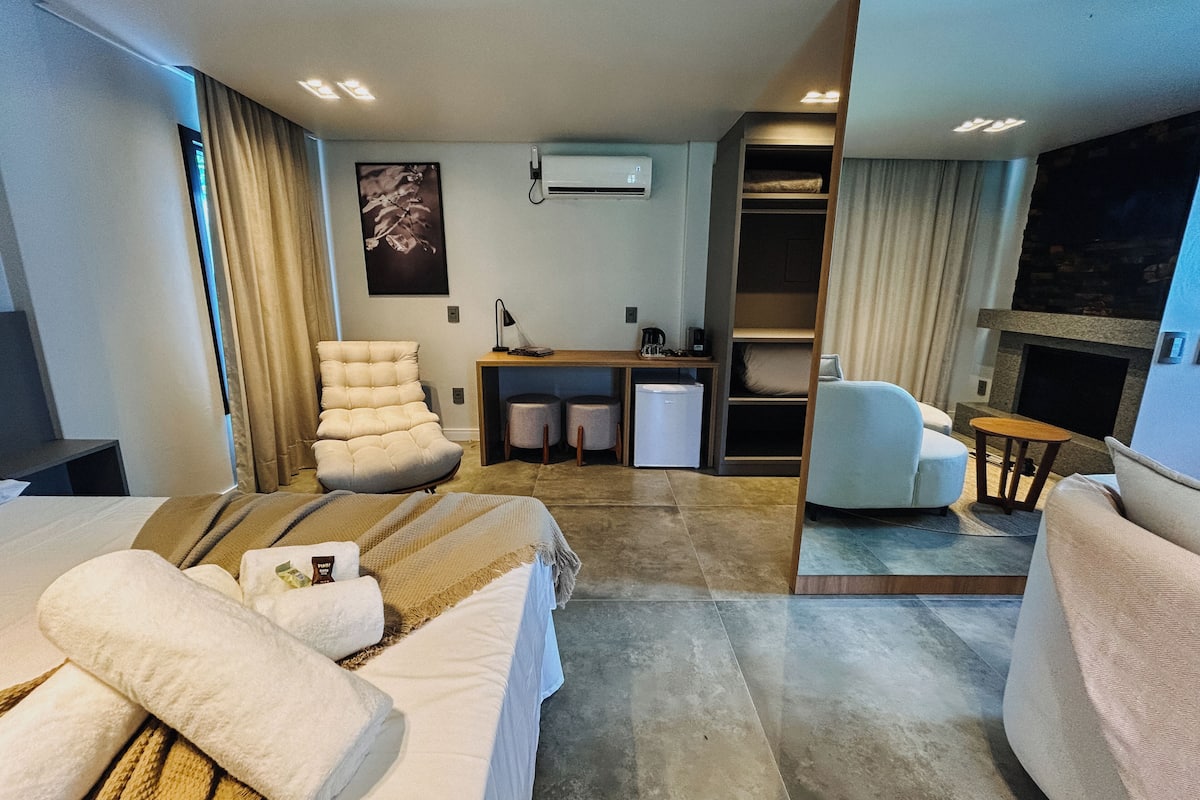 Quarto de hotel moderno com uma cama grande, área de estar com uma poltrona, pequena escrivaninha e lareira.