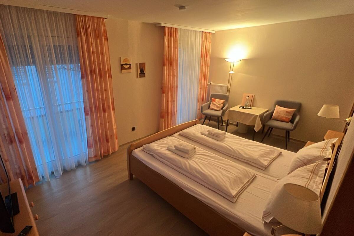 Quarto de hotel com uma cama grande, uma cadeira, uma mesinha e um abajur.