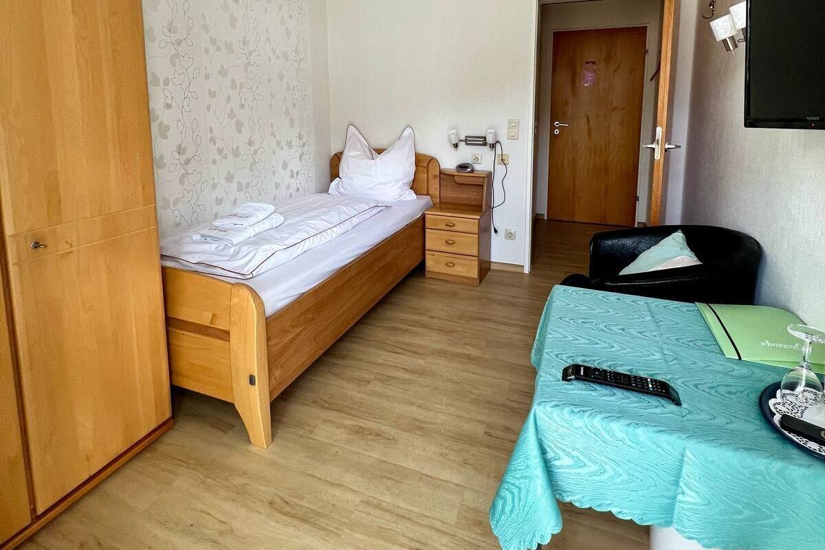 Um quarto pequeno e organizado com uma cama de solteiro, uma escrivaninha com cadeira, um guarda-roupa e uma televisão.