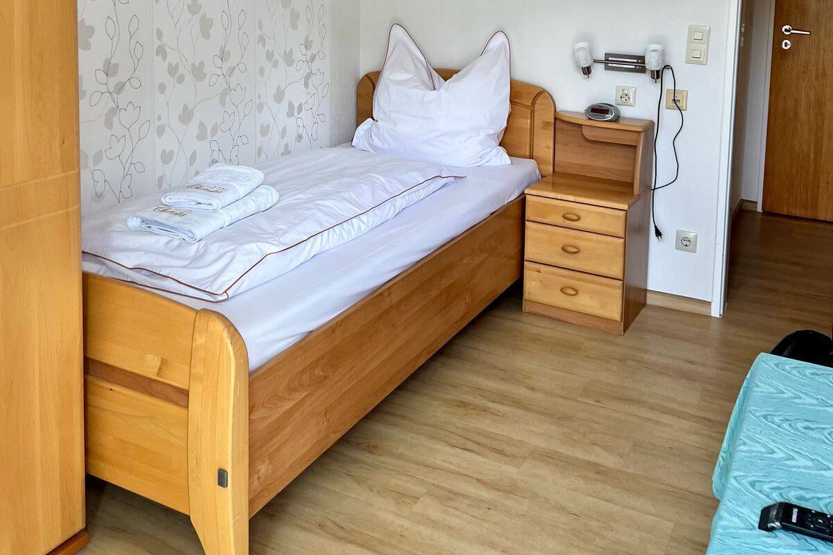 Quarto individual com uma cama de madeira, mesa de cabeceira, guarda-roupa e porta.