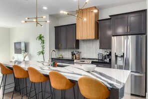Uma cozinha moderna com armários escuros, uma geladeira de aço inoxidável, uma ilha de mármore com banquetas e uma luminária pendente.