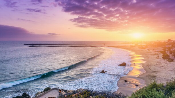 Vista costeira com praia, ondas e pôr do sol.