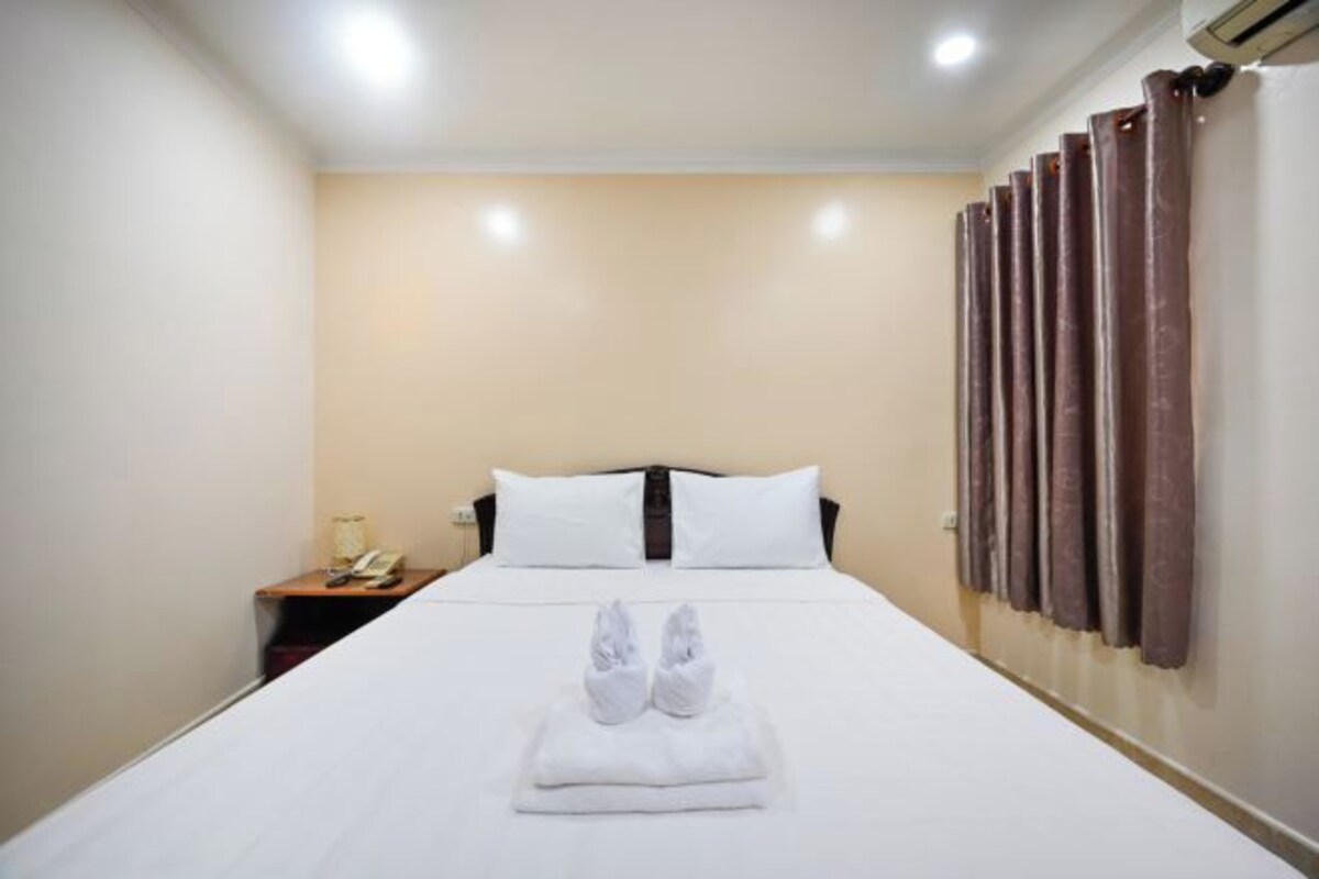 Quarto de hotel com cama bem arrumada, mesa de cabeceira com abajur e cortina na janela.