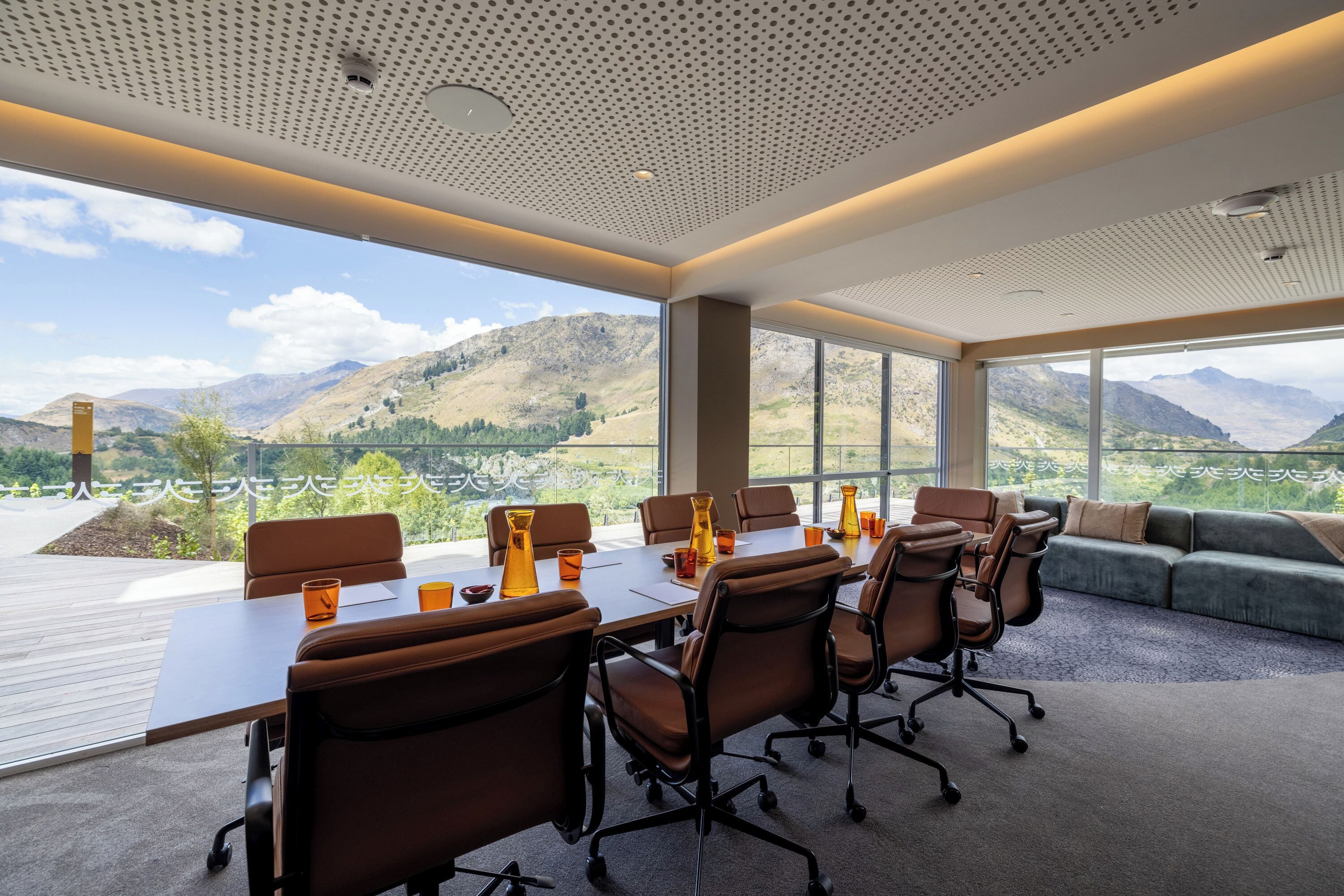 Uma sala de reuniões moderna com uma mesa grande e cadeiras, janelas amplas com vista para as montanhas e uma área de estar acolhedora.