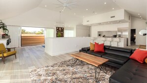 Uma sala de estar moderna com um sofá de couro preto, uma mesa de centro de madeira e um tapete estampado. Há uma área de cozinha com armários brancos e um ventilador de teto.