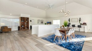 Uma sala de estar ampla com piso de madeira, uma mesa de jantar com cadeiras, uma estante de livros e um ventilador de teto.