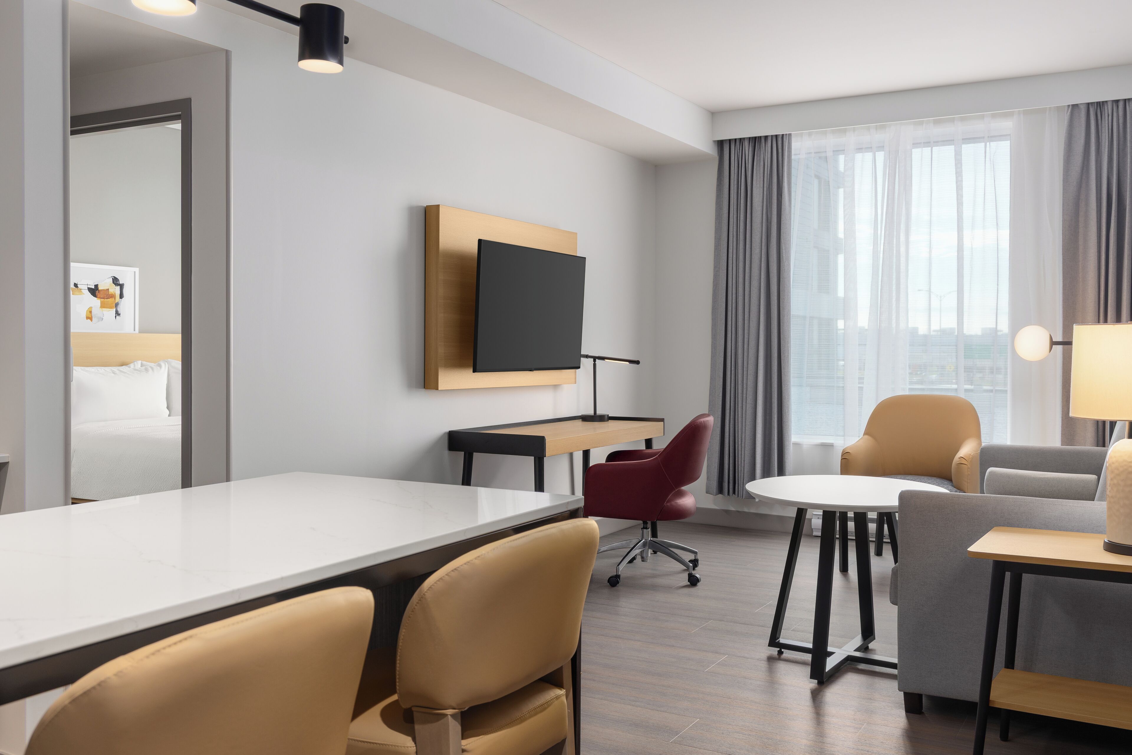 Quarto de hotel com cama, escrivaninha, televisão, cadeira, mesa e vista da cidade através de grandes janelas.