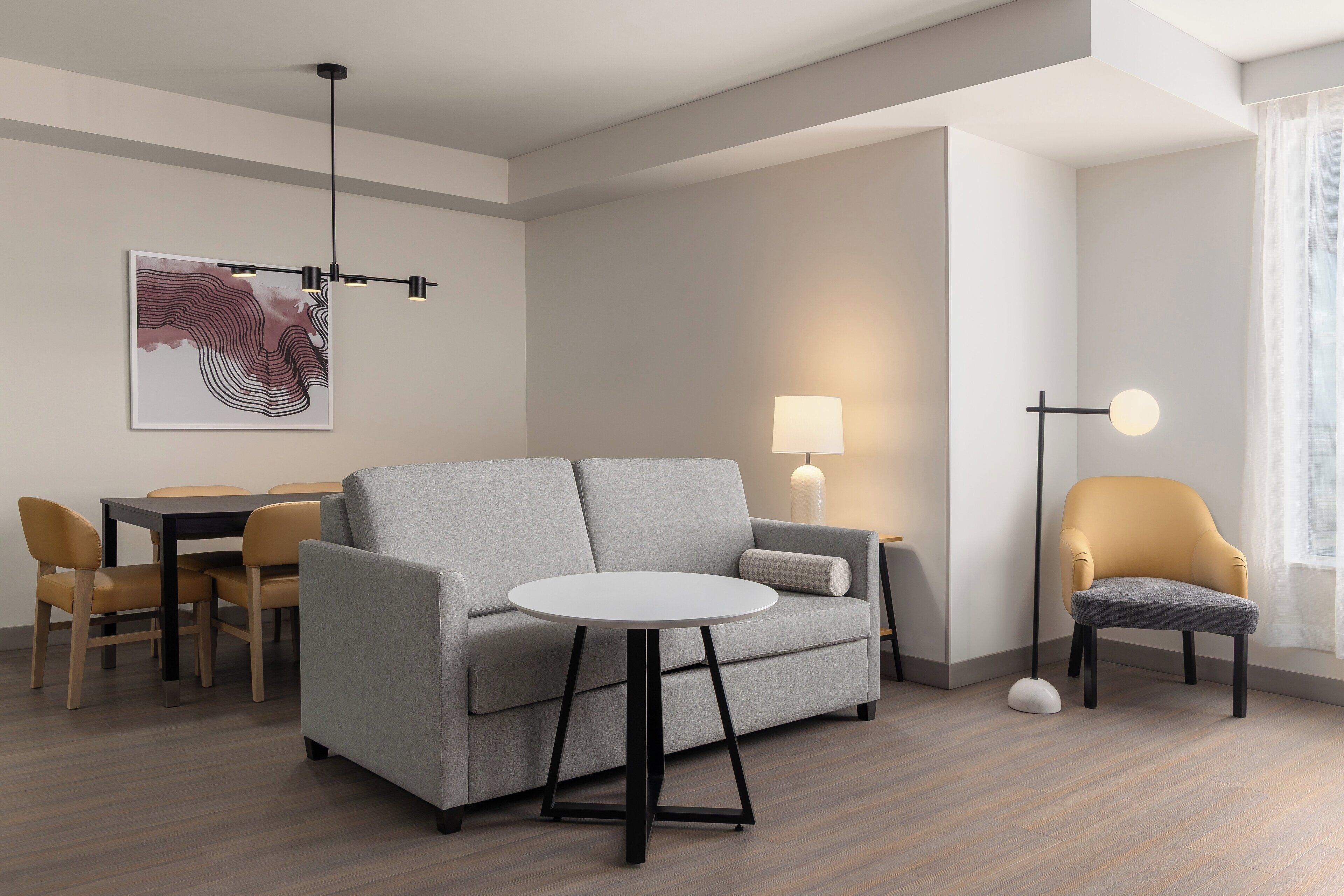 Uma sala de estar moderna com um sofá cinzento, uma mesa de centro redonda, uma mesa de jantar com cadeiras, um abajur de chão e um aplique na parede.