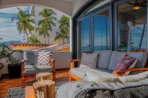 Varanda com sofá, cadeiras e uma mesa de madeira. Palmeiras e vista para uma praia com edifícios ao fundo.