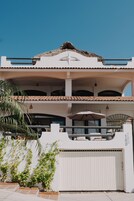 Um edifício de dois andares com fachada branca, telhado em telhas e varanda com mesa e guarda-sol.