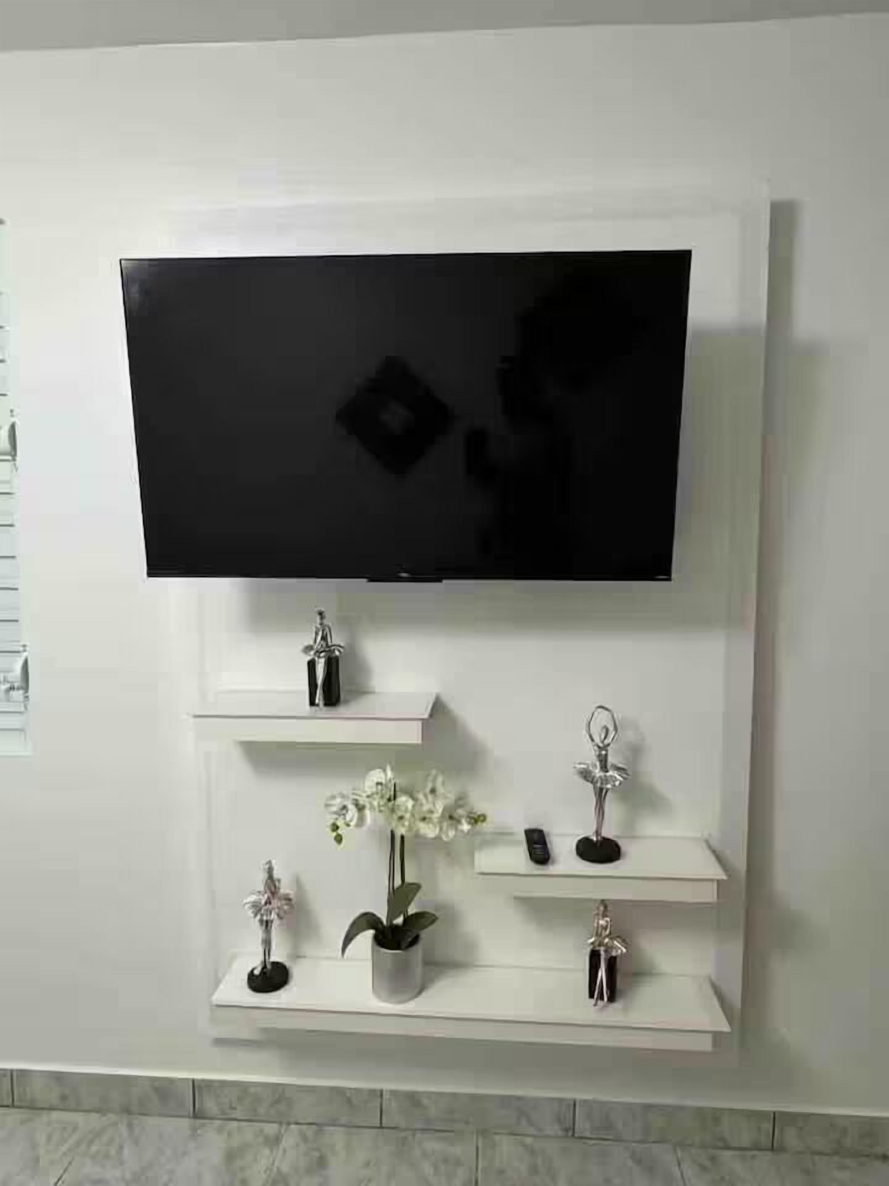 Uma sala de estar moderna com uma grande televisão de tela plana presa a uma parede branca. Abaixo da TV, há três prateleiras brancas com objetos decorativos, incluindo uma planta em vaso e pequenas esculturas.