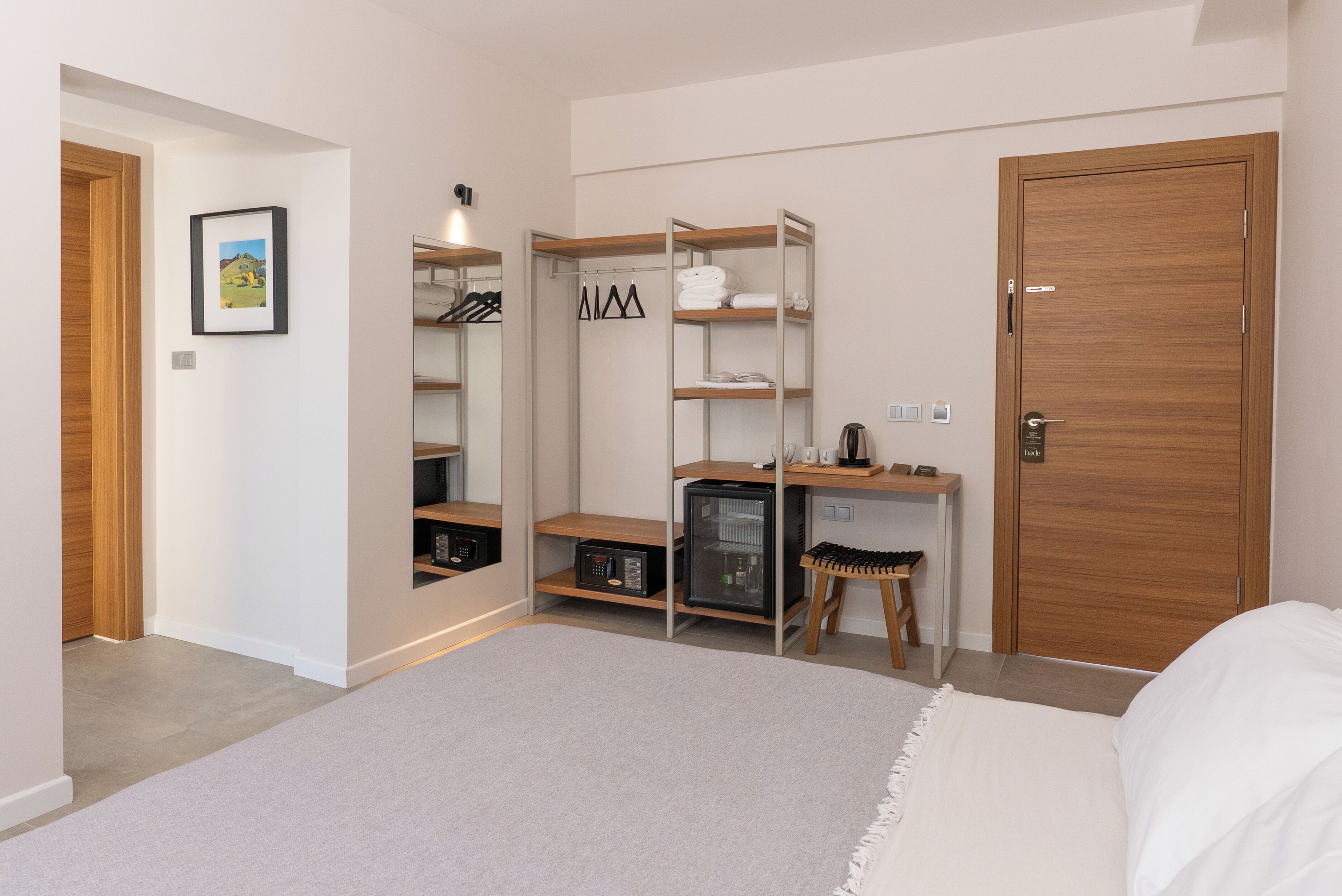 Quarto com cama, guarda-roupa de madeira, escrivaninha com cadeira e estante com roupas dobradas e um pequeno frigorífico.