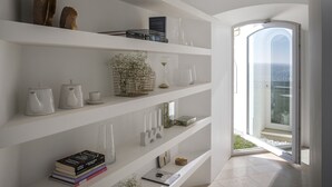 Um interior moderno com estantes brancas exibindo livros, objetos decorativos e vista do exterior por uma porta de vidro.