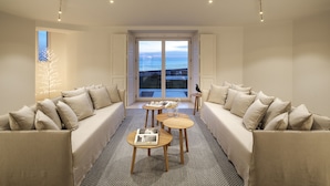 Uma sala de estar moderna com um sofá, duas mesas redondas de madeira e uma grande porta de vidro deslizante que dá para uma varanda com vista para o mar.