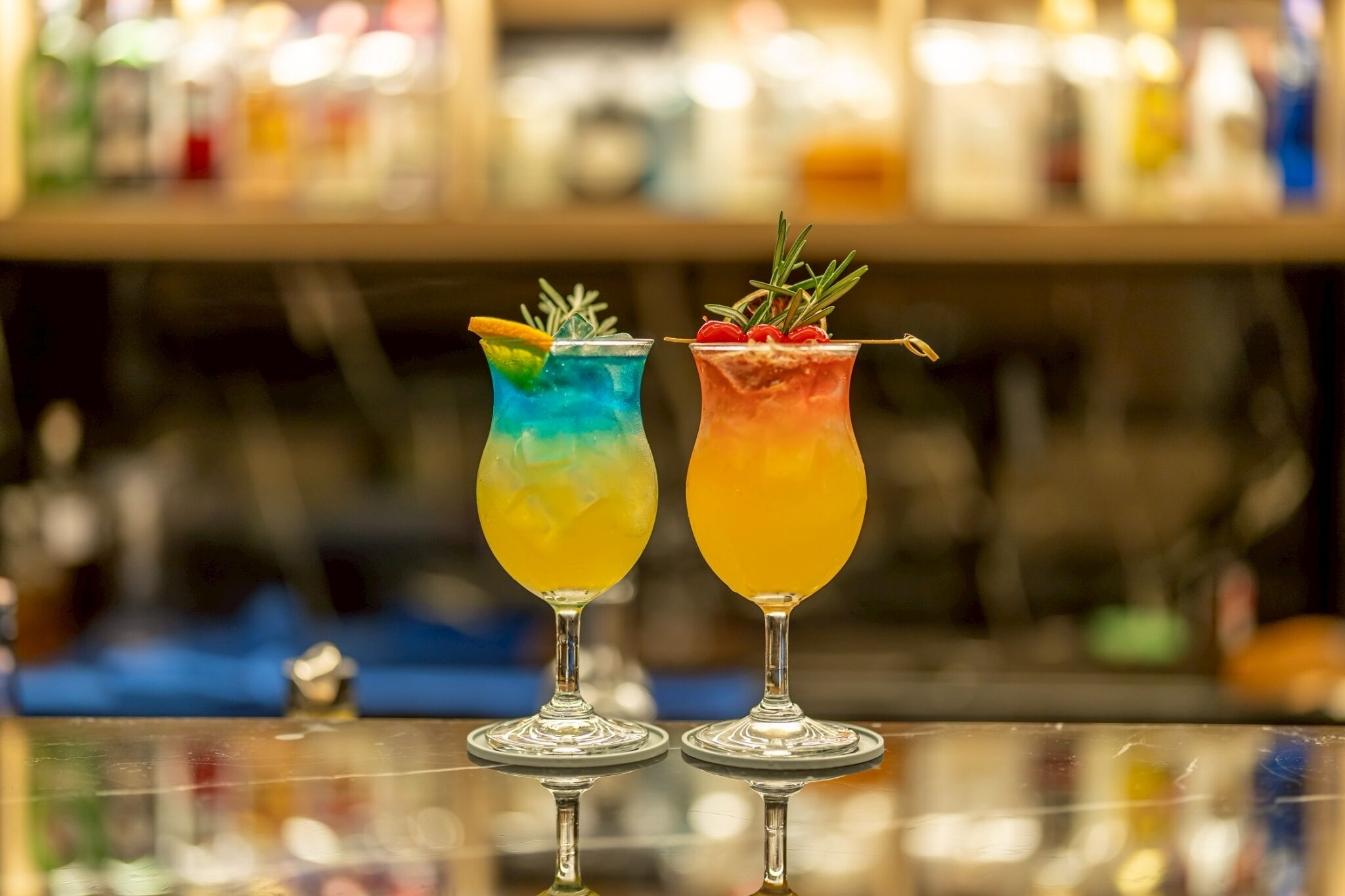 Dois coquetéis coloridos em um balcão de bar, com um fundo desfocado de prateleiras.