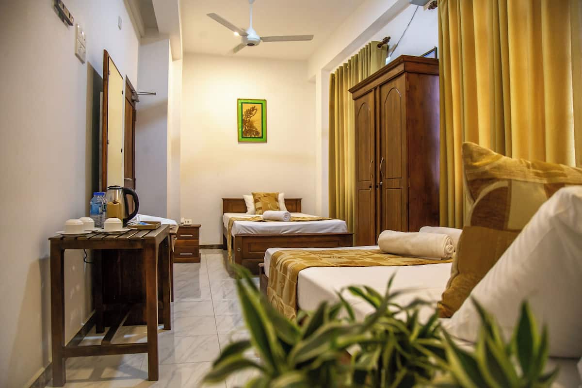 Quarto de hotel com duas camas, um guarda-roupa de madeira, uma mesa com chaleira e um ventilador de teto.