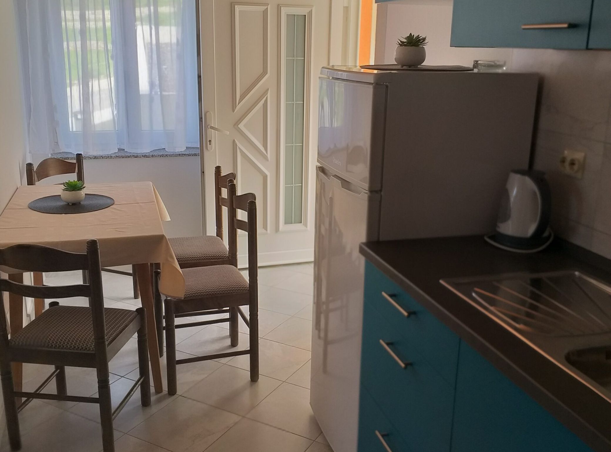 Cozinha compacta com geladeira, pia e fogão. Há uma mesa de jantar com cadeiras e um vaso de planta.