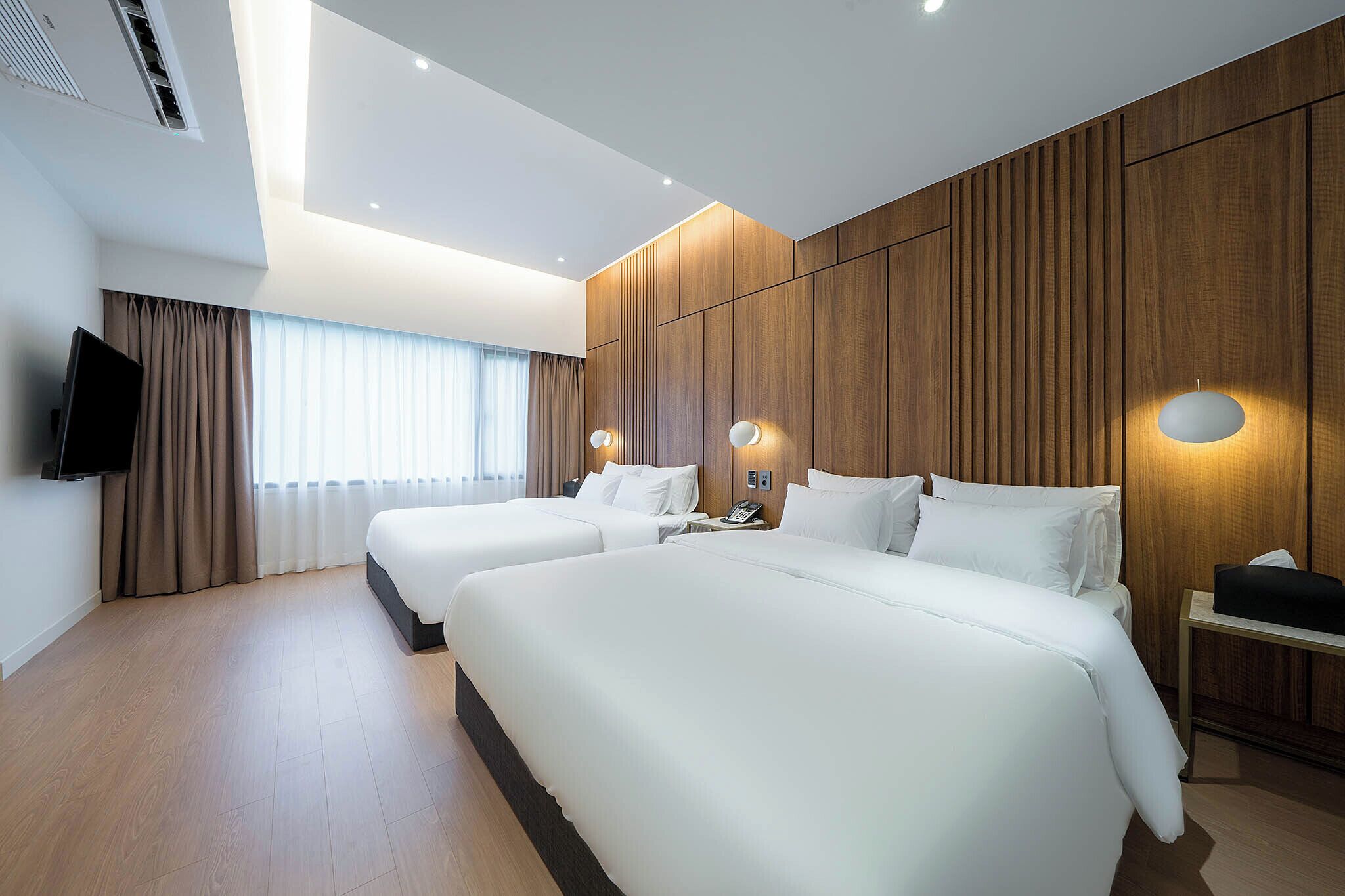 Quarto de hotel com duas camas, painéis de madeira nas paredes, televisão de tela plana na parede e uma janela com cortinas.