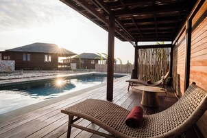 Um deck de madeira com piscina, cadeiras de vime e área coberta com vista para casas ao fundo.