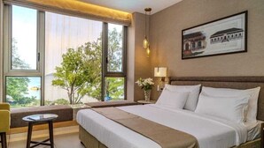 Quarto de hotel com uma cama grande, uma mesinha de cabeceira, uma cadeira, uma janela com vista para árvores e um quadro na parede.
