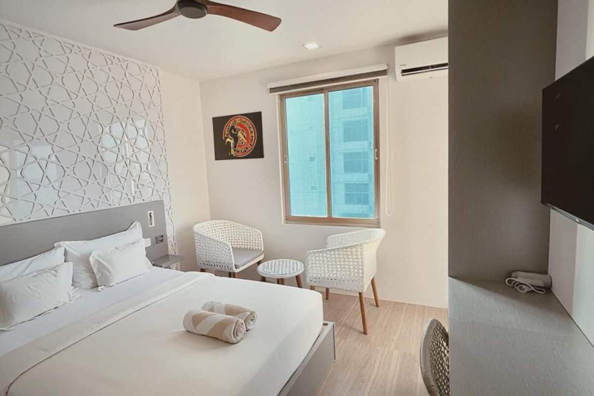 Quarto de hotel com cama, ventilador de teto, televisão, janela com vista e uma cadeira.
