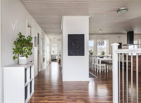 Um corredor espaçoso com soalho de madeira, paredes brancas e um pilar central. Há uma área de jantar com cadeiras brancas e uma mesa, e uma planta em vaso à esquerda.