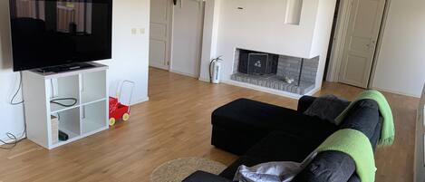 Uma sala de estar com uma TV de tela plana sobre um suporte branco, um sofá preto com um cobertor verde e uma lareira.