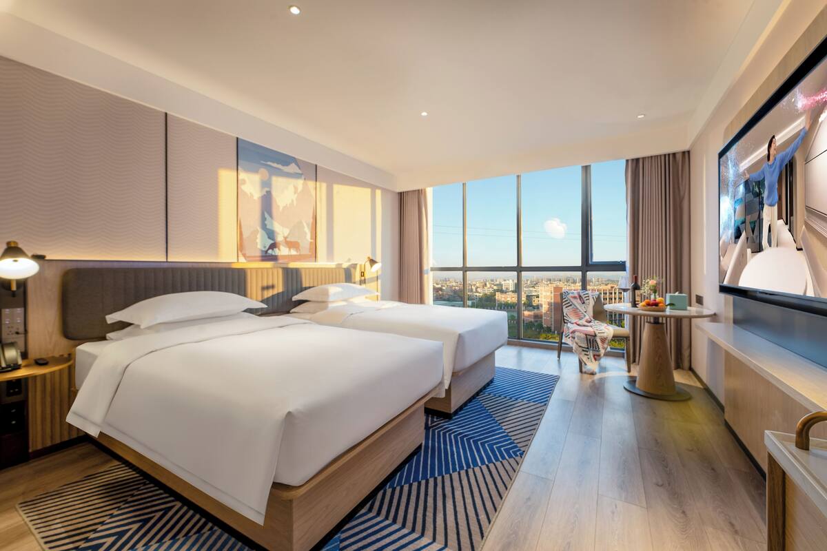 Quarto de hotel com duas camas, uma janela grande com vista para a cidade, uma mesa com cadeira e uma televisão.