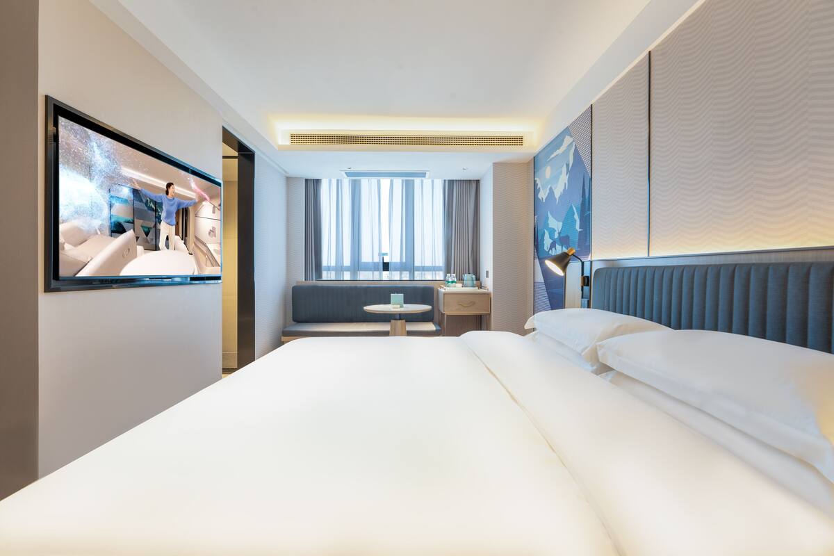 Quarto de hotel moderno com uma cama grande, área de estar, escrivaninha e televisão.