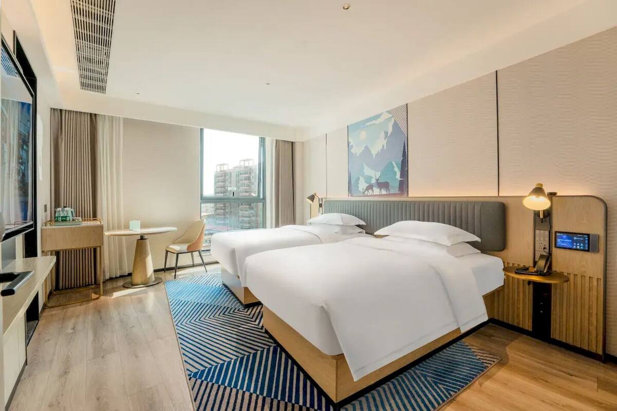 Quarto de hotel com duas camas, uma escrivaninha, uma cadeira, televisão e janela com vista para a cidade.