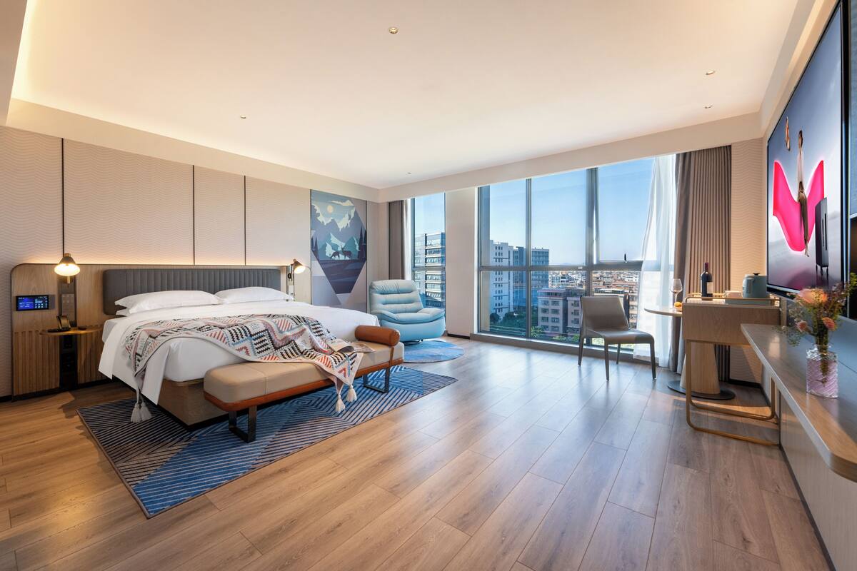Quarto de hotel moderno com uma cama grande, uma escrivaninha, uma cadeira e vista para a cidade.