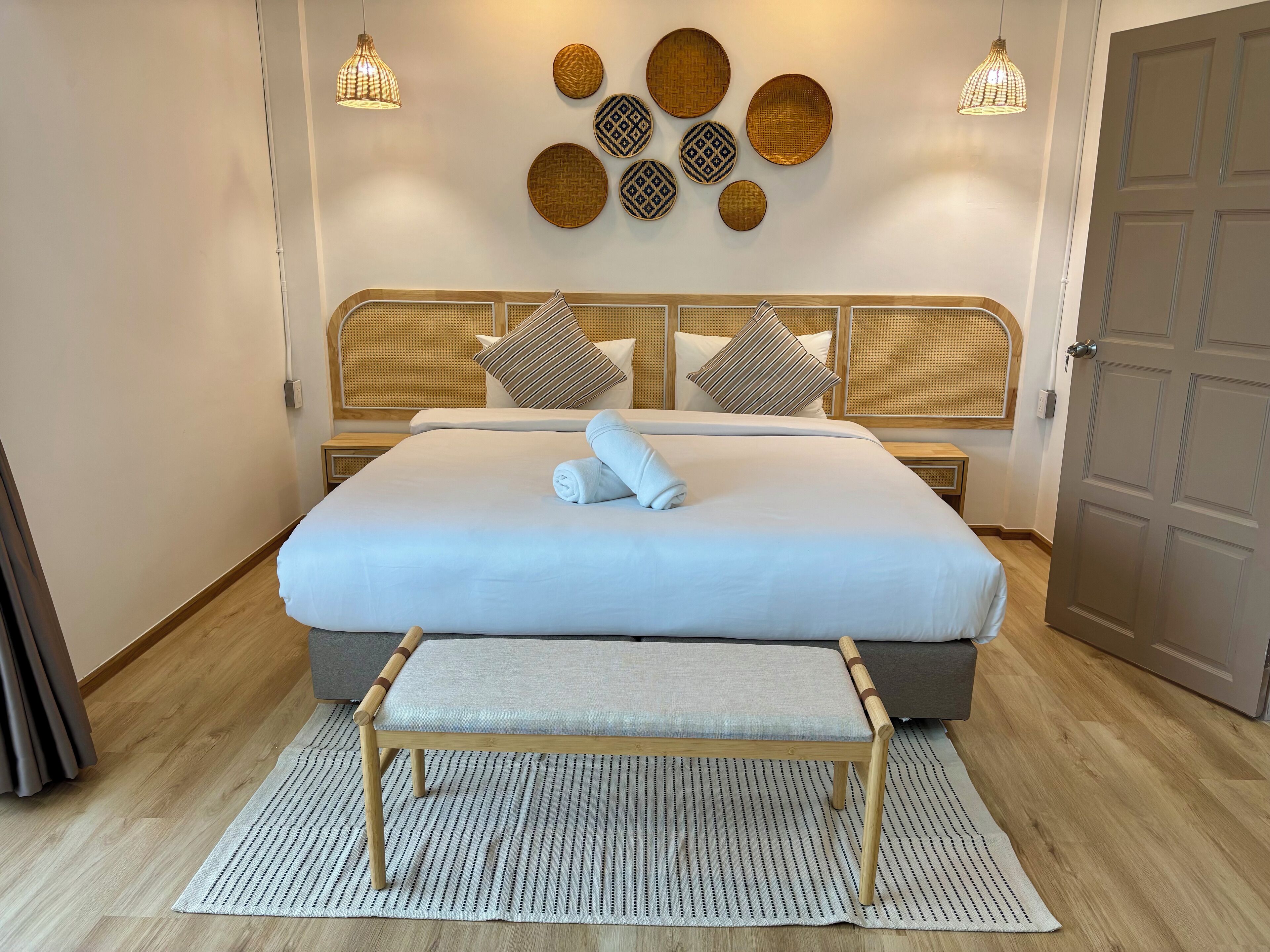 Uma cama bem arrumada com edredom branco e duas almofadas, um banco de madeira ao pé da cama, cabeceira trançada e um painel com adornos circulares na parede.