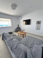 Uma sala de estar moderna com um sofá cinzento grande, uma mesa de centro de madeira, uma TV montada na parede e um quadro com uma palmeira.