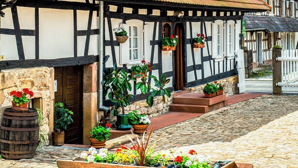 Uma casa tradicional de enxaimel com um pátio de calçamento, um vaso de madeira com flores e cestos de flores pendurados.