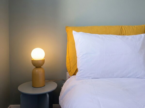 Uma cama com roupa de cama branca e um travesseiro amarelo, uma mesa redonda com uma luminária, e uma parede lisa.