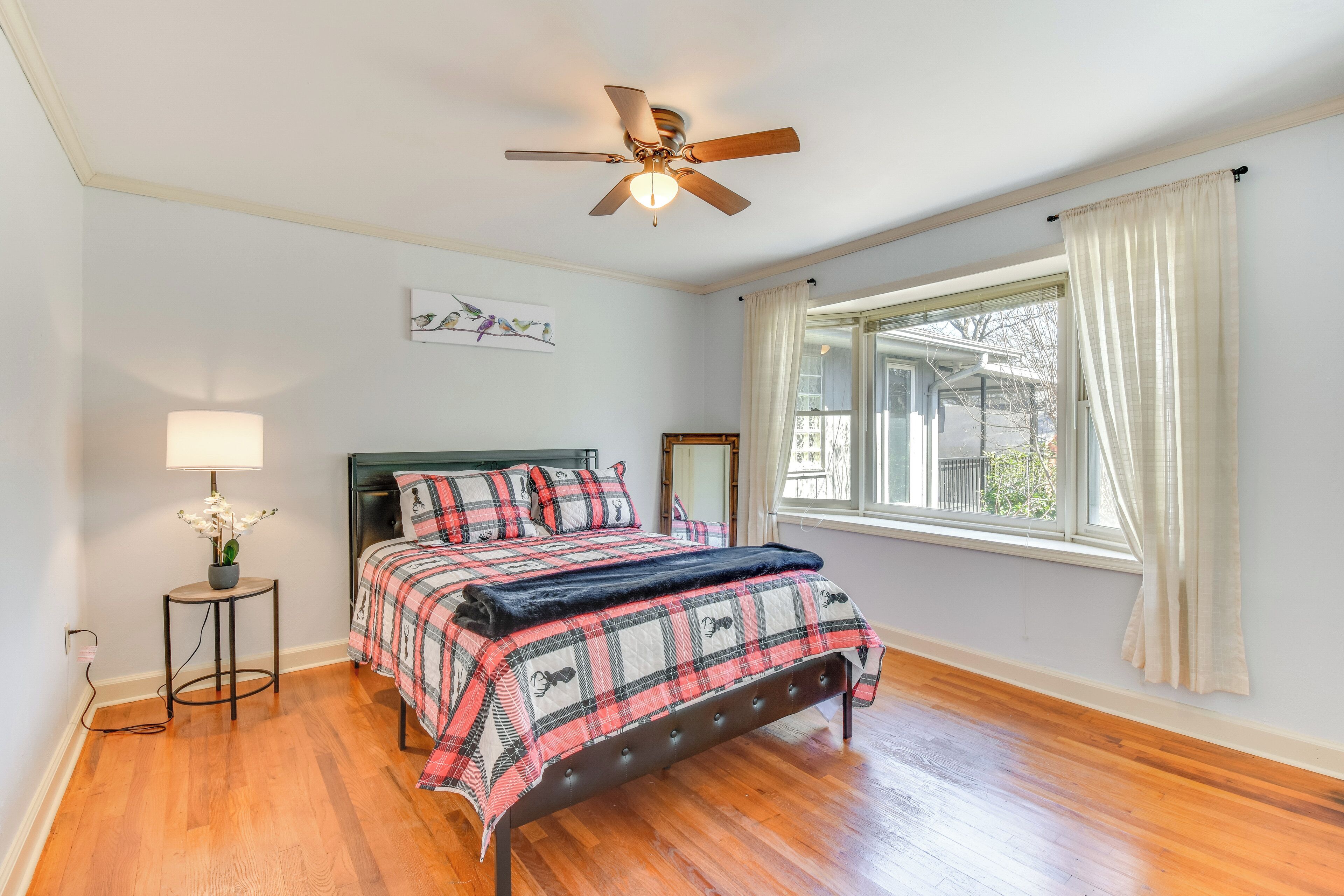 Quarto com cama, ventilador de teto, janela com cortinas, mesa de cabeceira com abajur e uma pequena planta.