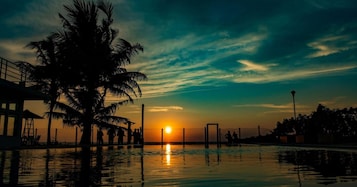 Pôr do sol sobre uma piscina, com palmeiras e uma cerca em primeiro plano.