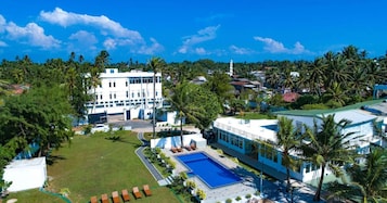 Um edifício branco grande com piscina e espreguiçadeiras, rodeado por palmeiras e vegetação.