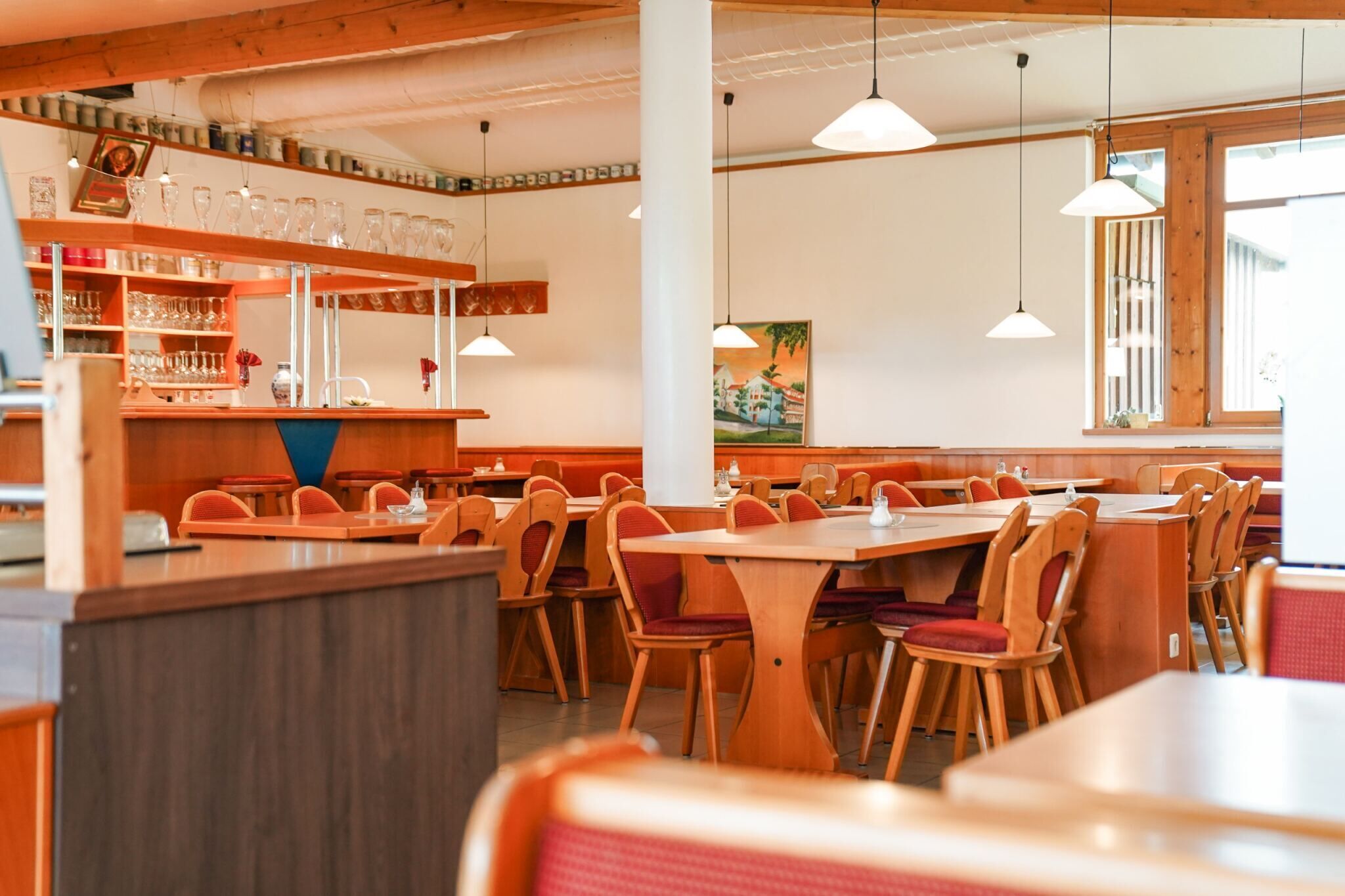 Interior de restaurante com mesas e cadeiras de madeira, área de bar com prateleiras cheias de copos, e iluminação pendente.